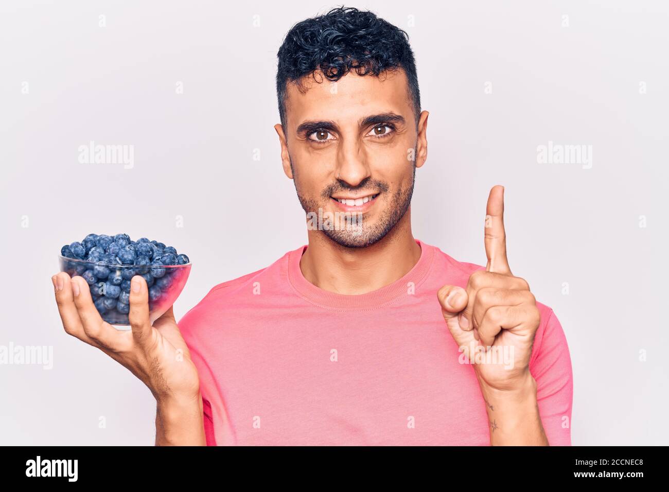 Young hispanic man holding blueberries smiling with an idea or question ...