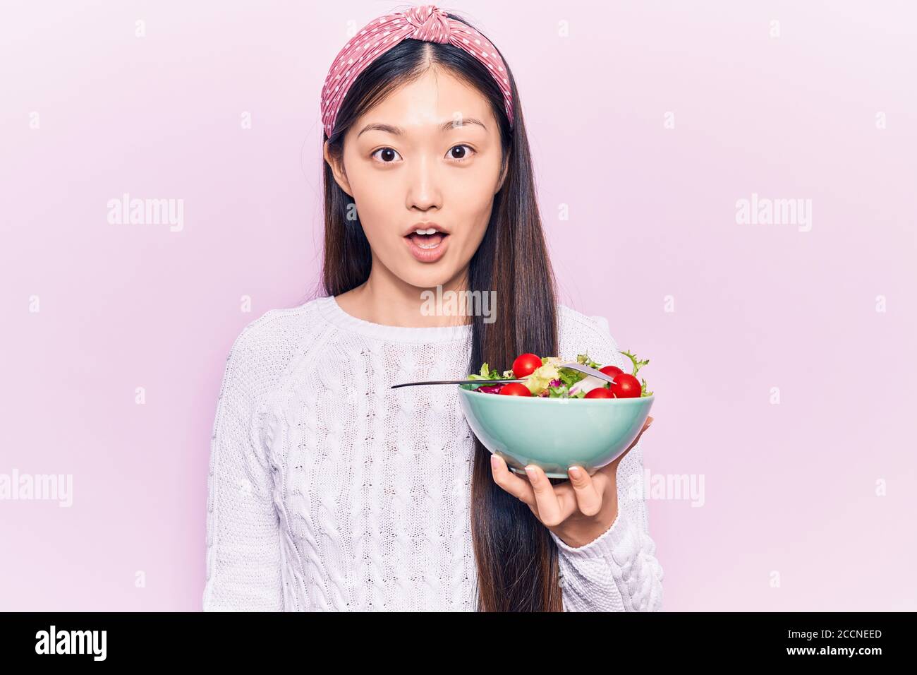 Young beautiful chinese woman eating bowl with salad scared and amazed ...