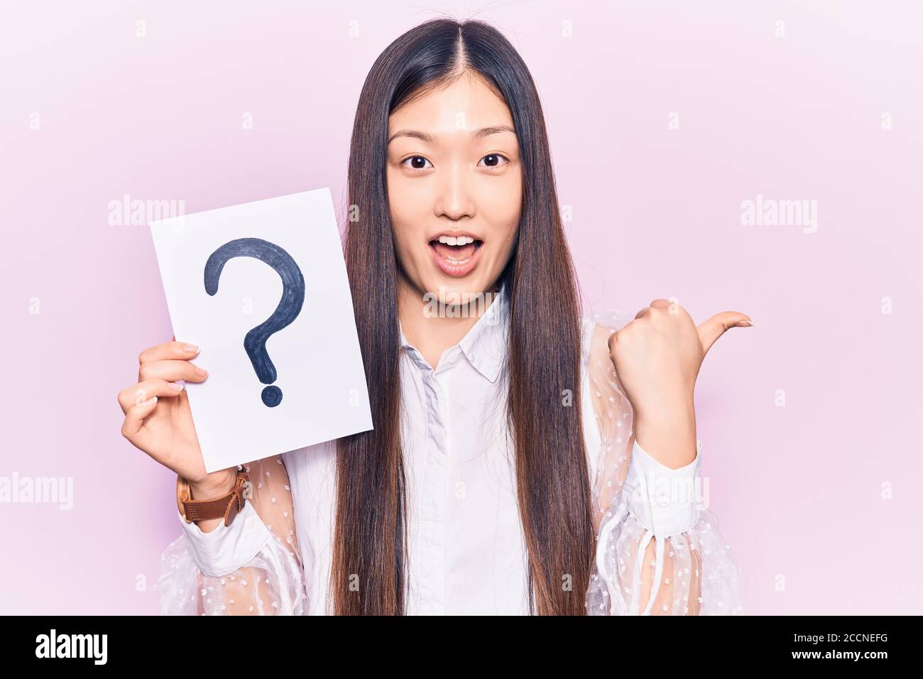 Young beautiful chinese woman holding question mark pointing thumb up ...