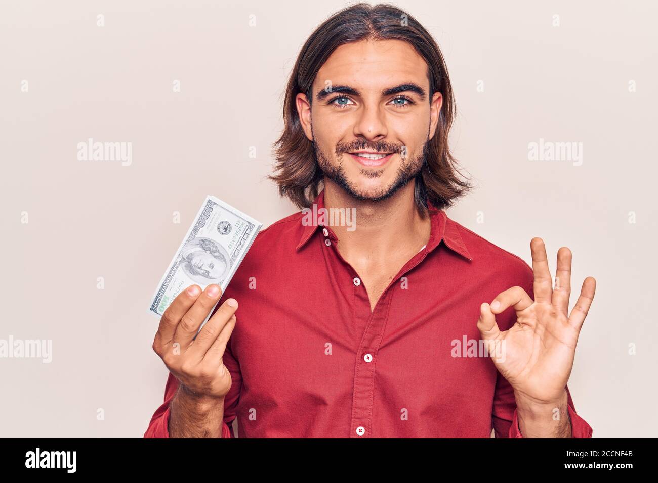 Young handsome man holding dollars doing ok sign with fingers, smiling ...