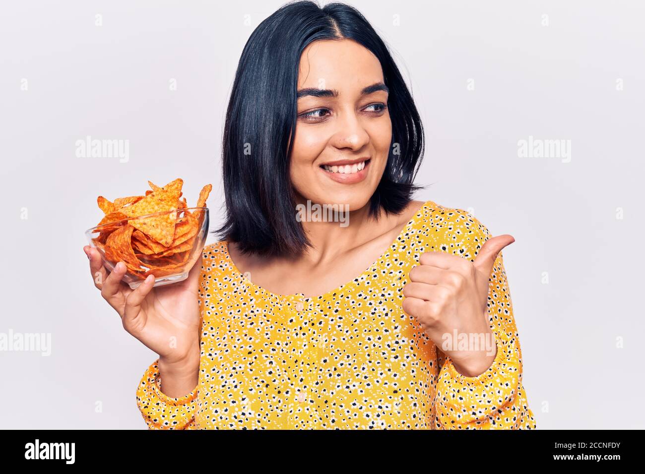 Young beautiful latin woman holding nachos potato chips pointing thumb ...