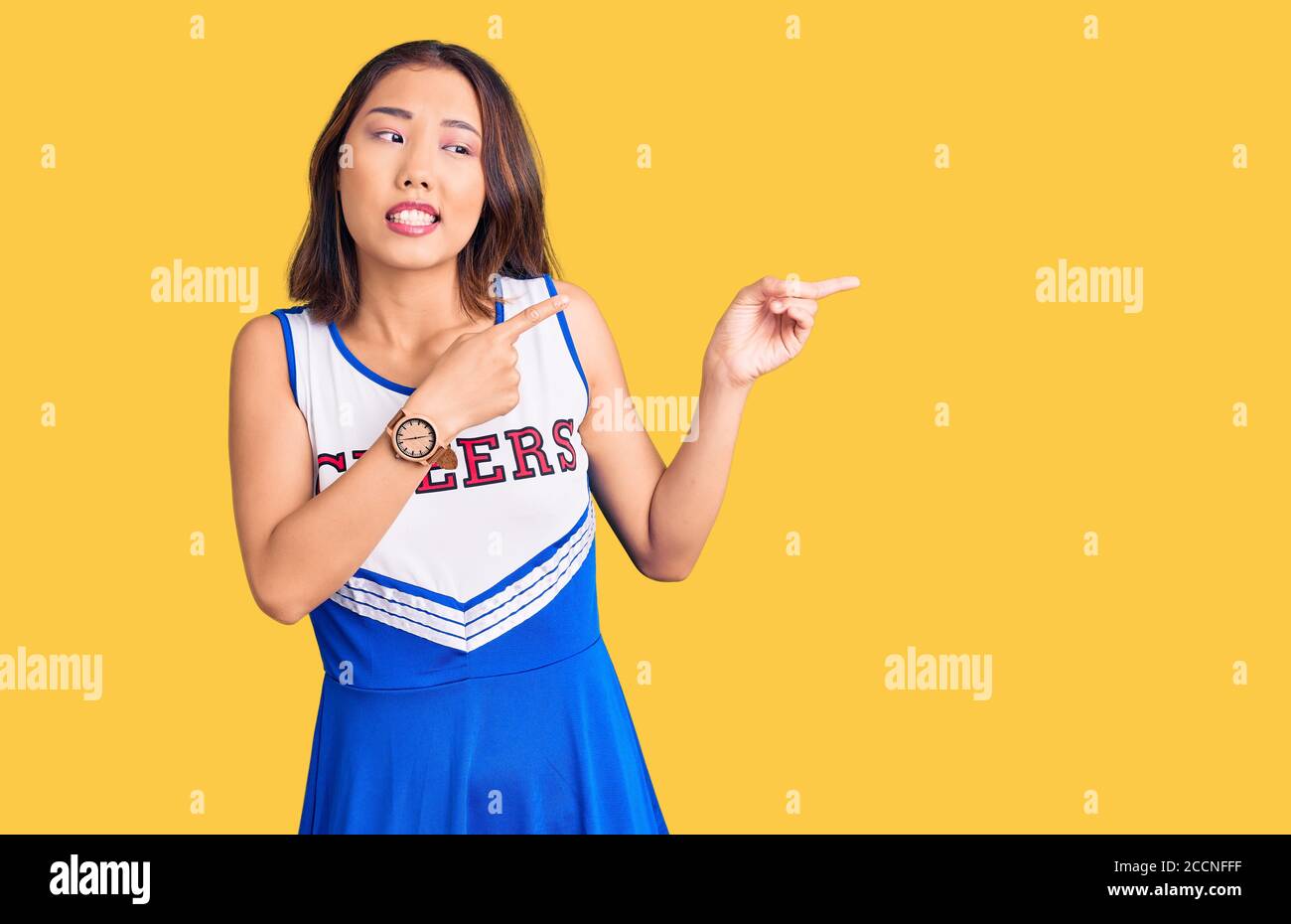 Young beautiful chinese girl wearing cheerleader uniform pointing aside ...