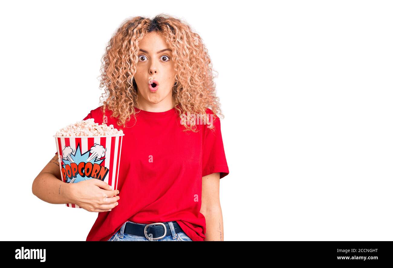Young blonde woman with curly hair eating popcorn scared and amazed ...