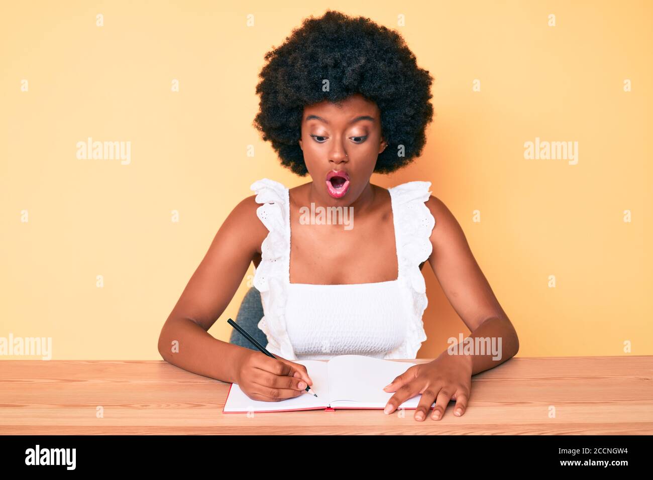 Young african american woman writing book scared and amazed with open ...
