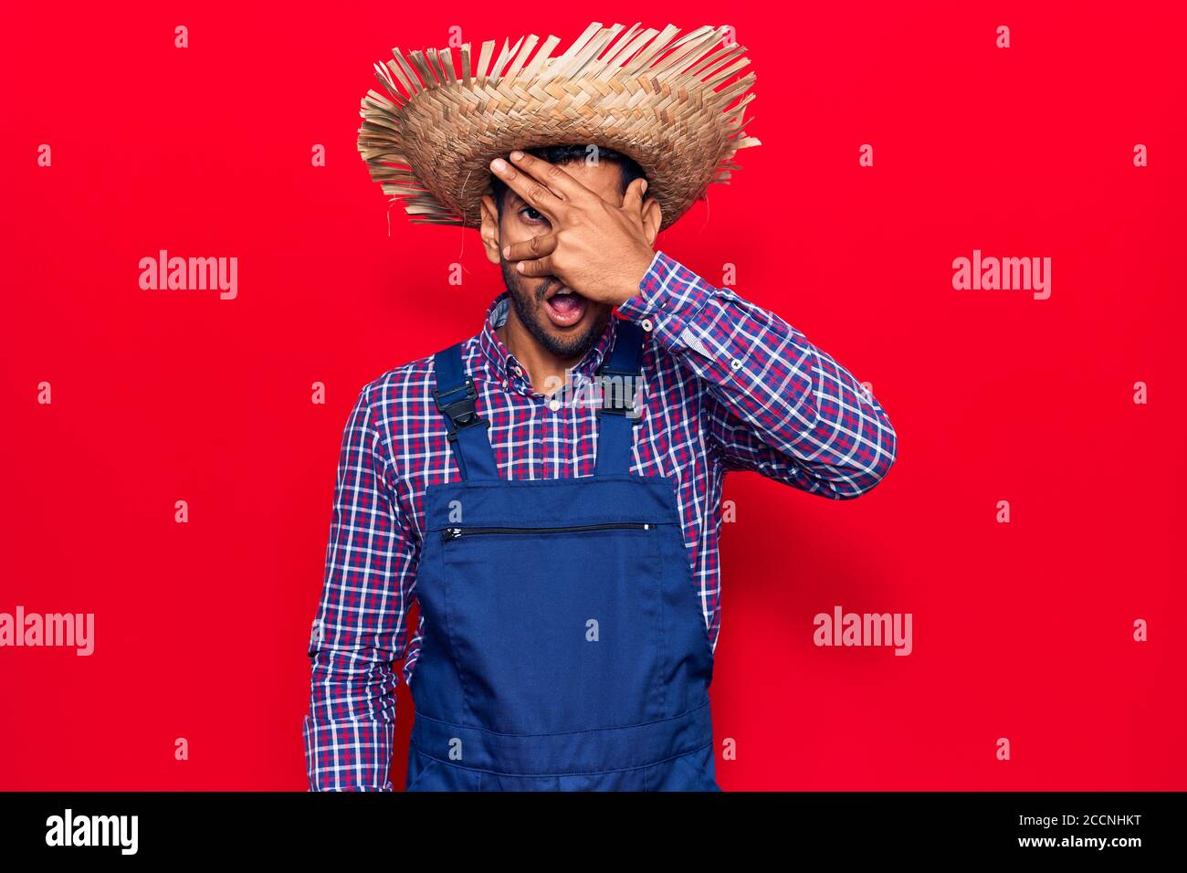 Young latin man wearing farmer hat and apron peeking in shock covering ...