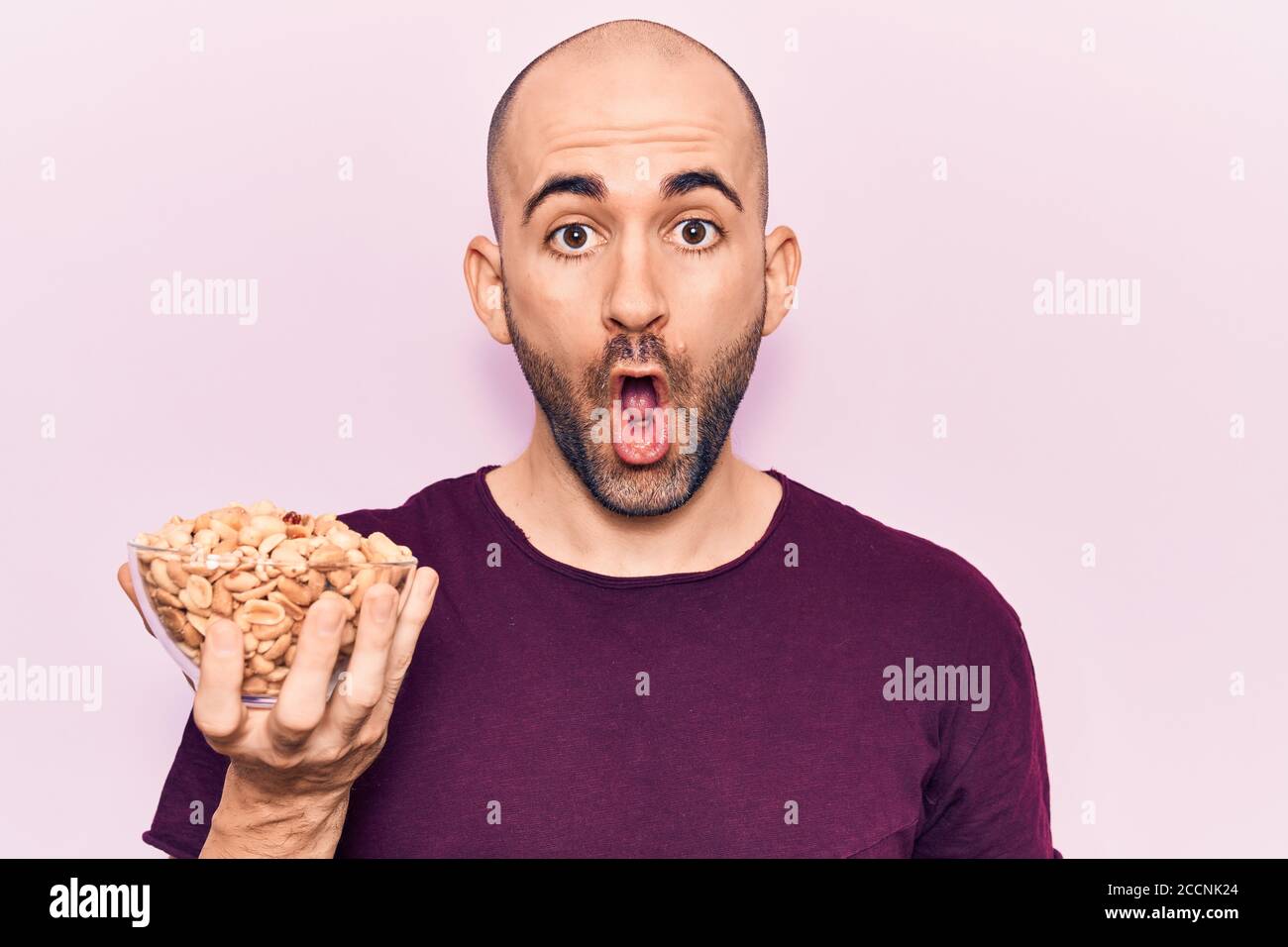 Young handsome bald man holding bowl with peanuts scared and amazed ...