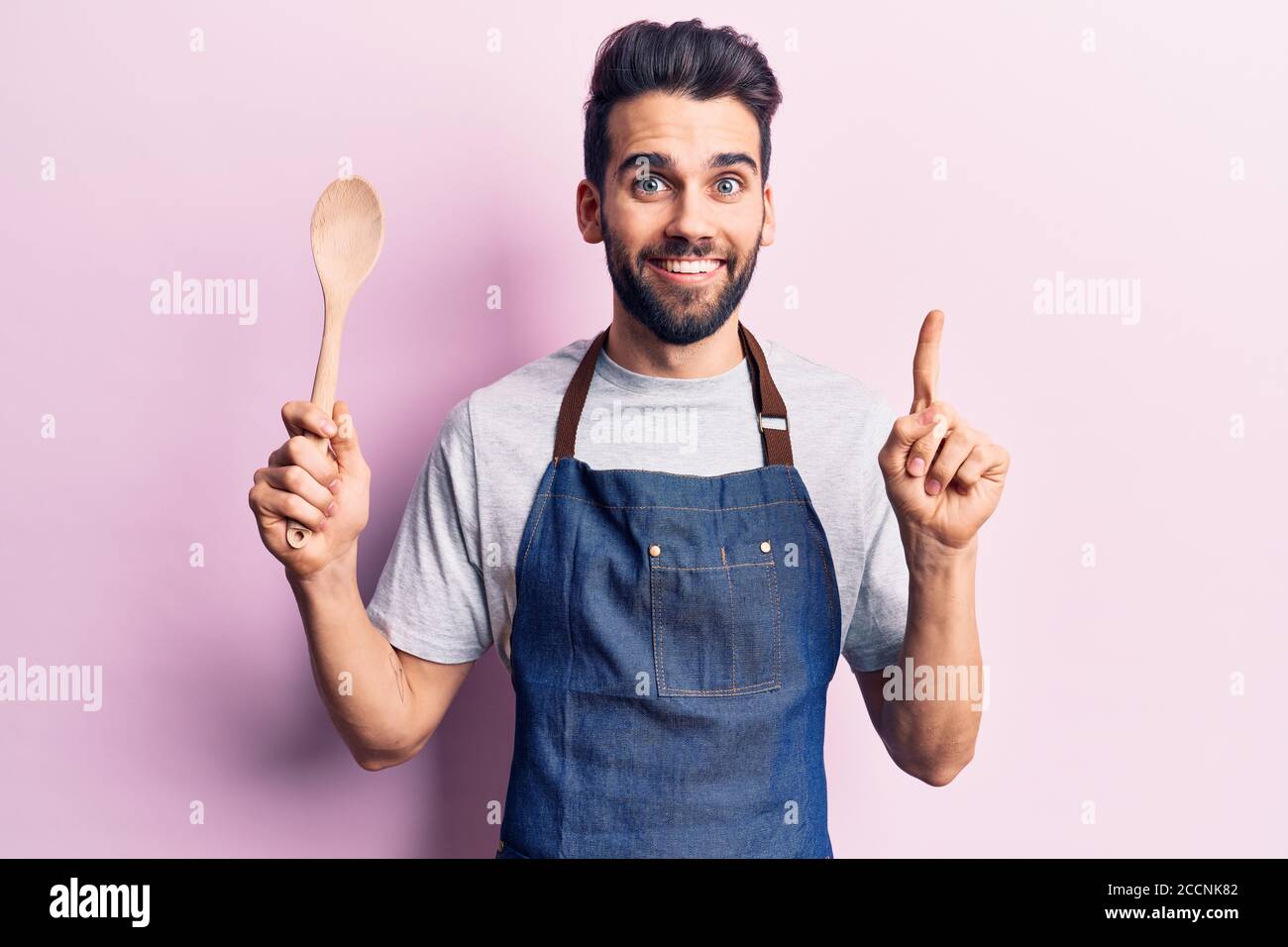 Young handsome man with beard wearing apron holding wooden spoon ...