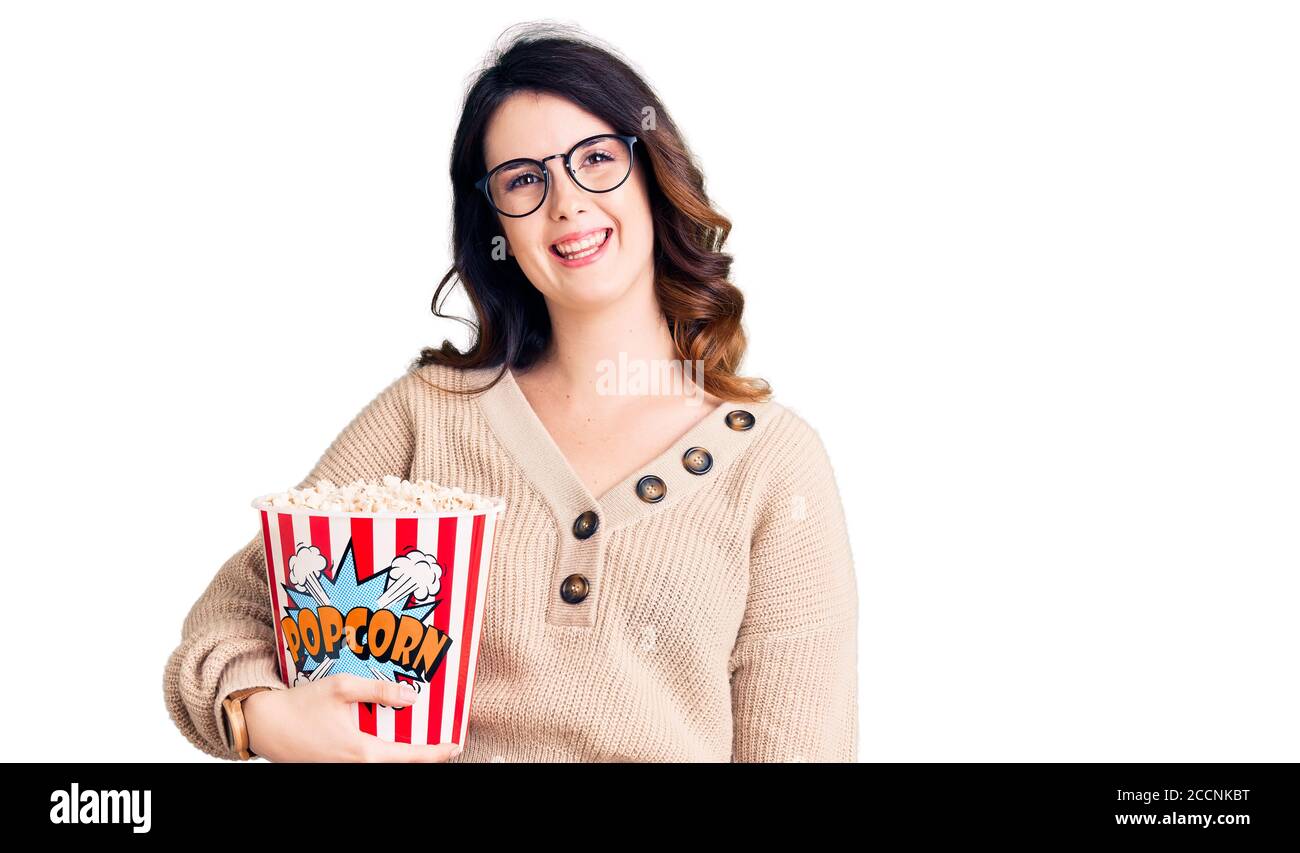 Beautiful young brunette woman eating popcorn looking positive and ...