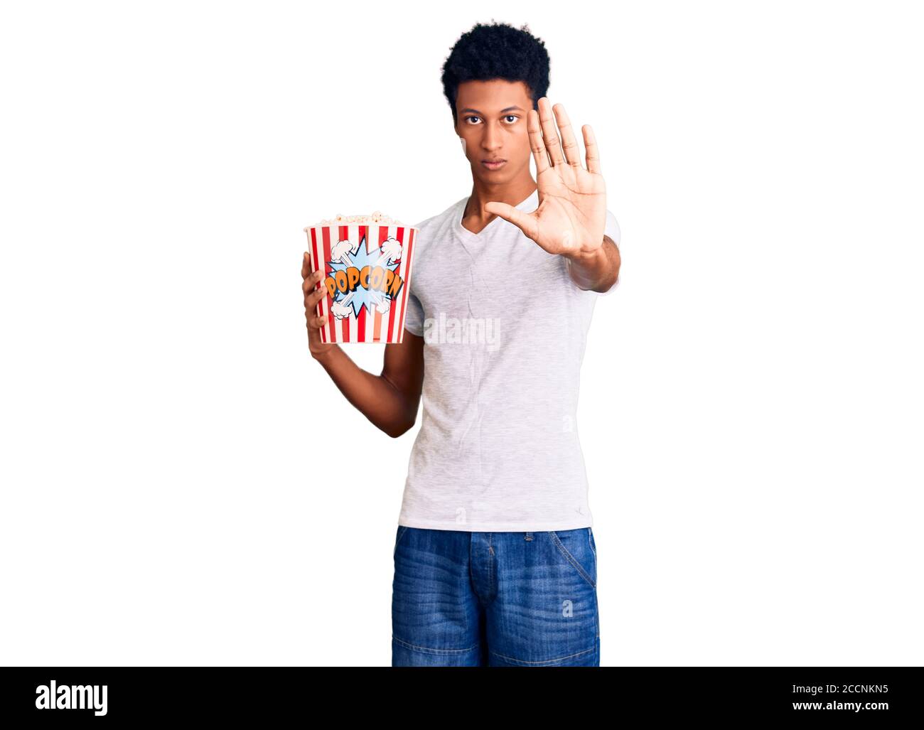 Young african american man holding popcorn with open hand doing stop sign with serious and ...