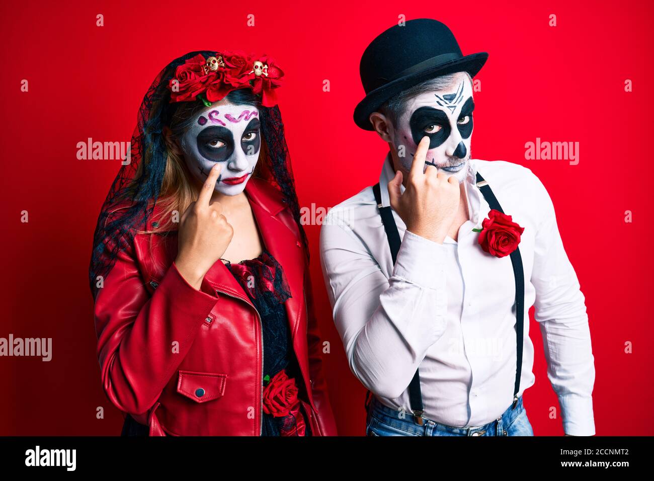 Couple wearing day of the dead costume over red pointing to the eye ...