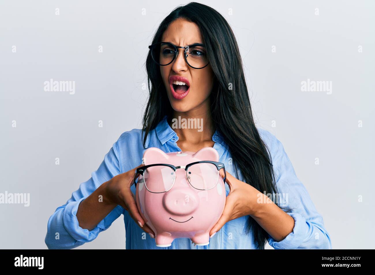 Beautiful hispanic woman holding piggy bank with glasses angry and mad ...