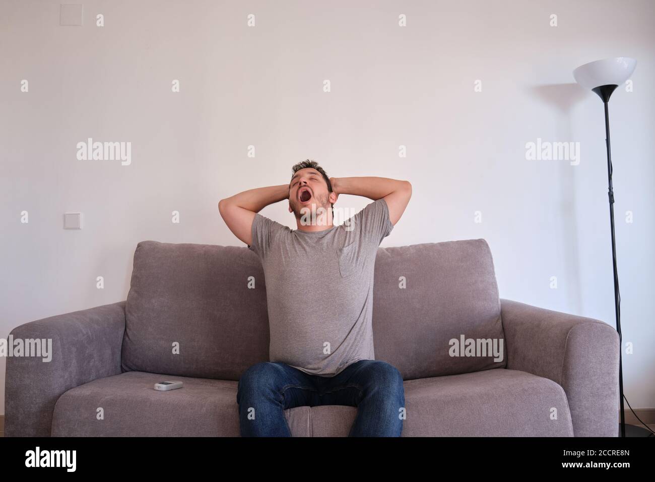 Portrait of a bored and tired young man sitting on a sofa yawning and ...