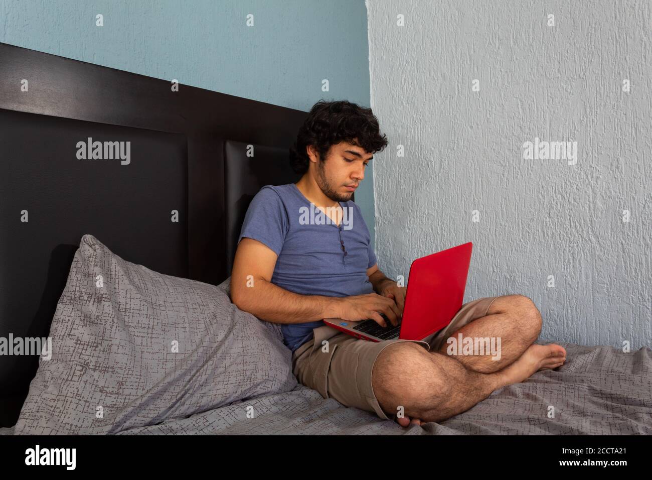 Hispanic young man using a laptop on a bed to work from home or study ...