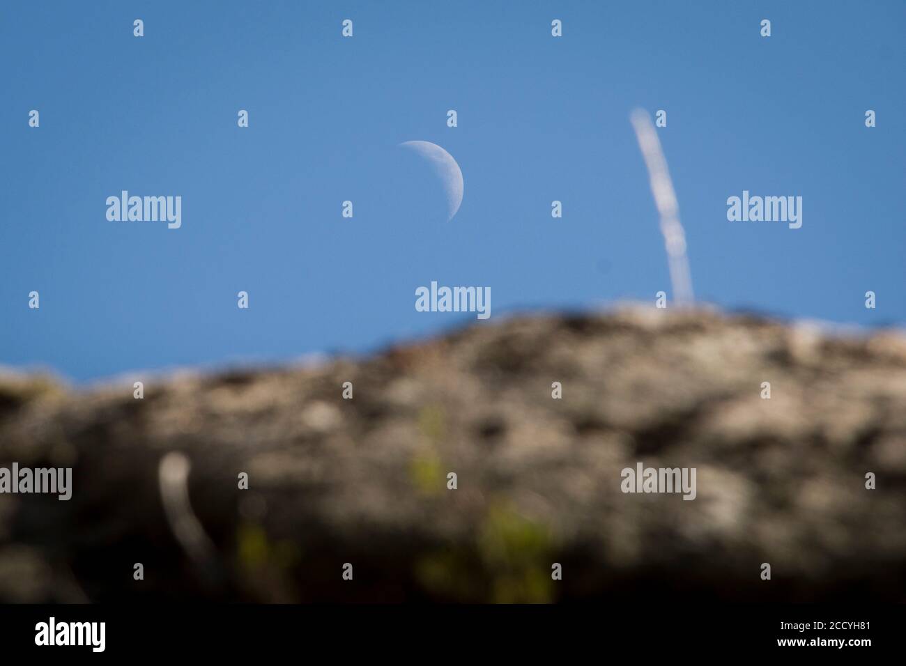 A crescent moon rises behind landscape rocks and vegetation during the ...