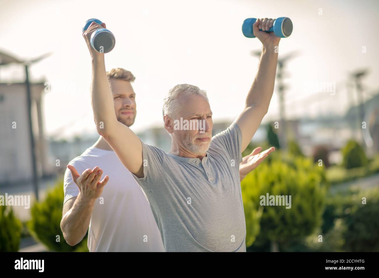 Grey-haired man raising hands with dumbbells, bearded instructor ...