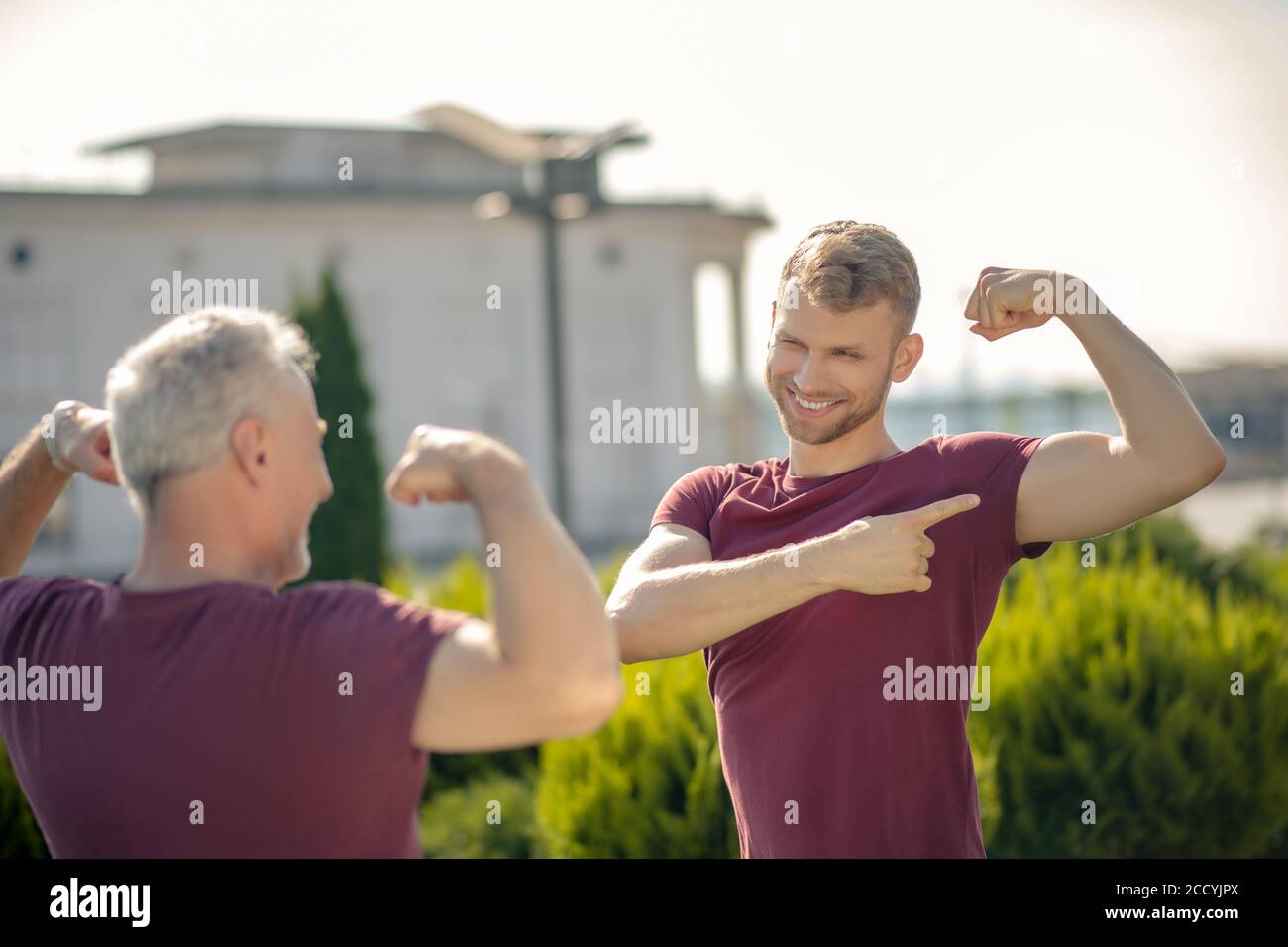 Bearded male showing biceps to grey-haired man, smiling Stock Photo - Alamy