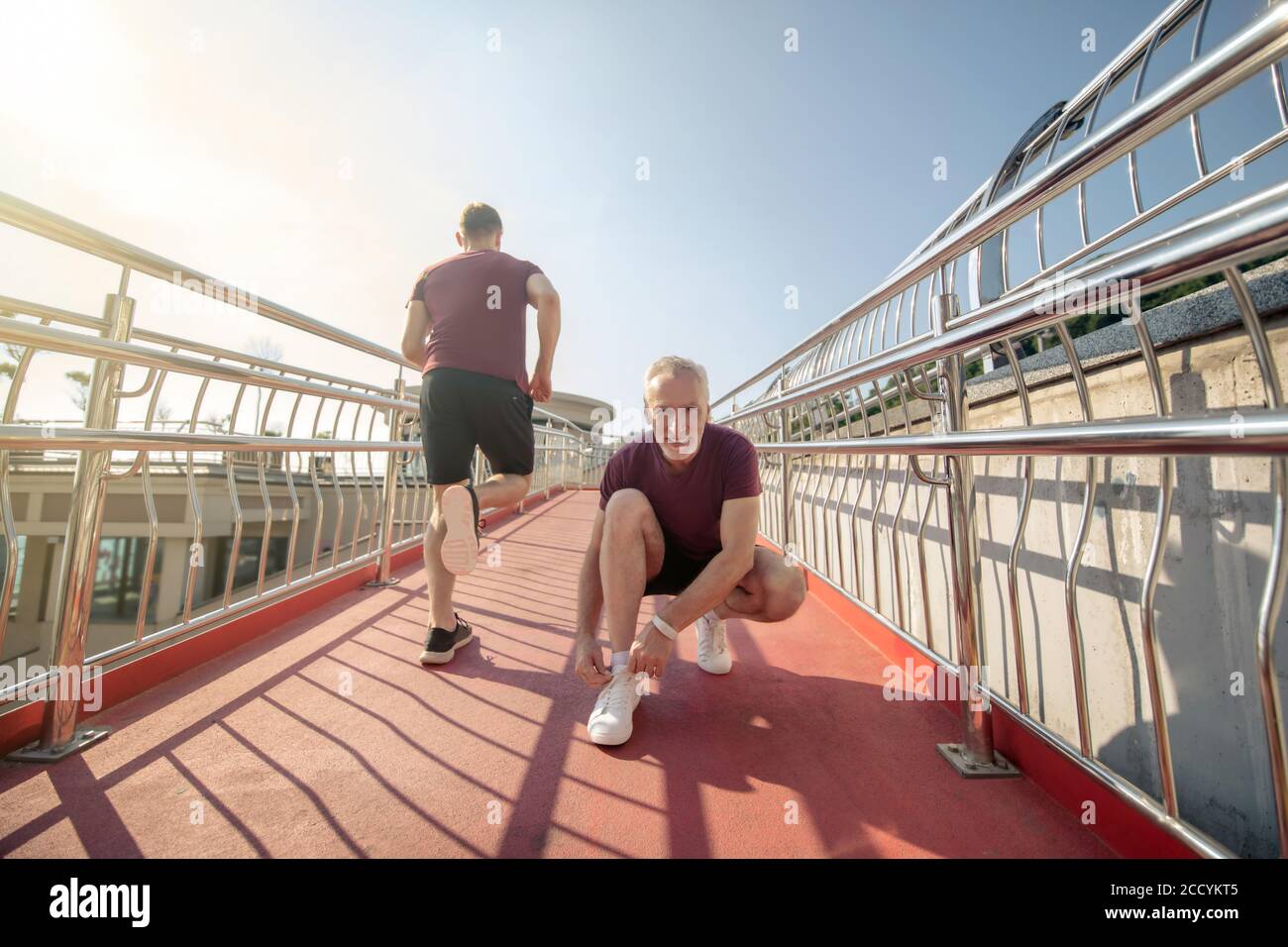 Grey-haired male crouching, lacing up his sneakers, yound male running ...