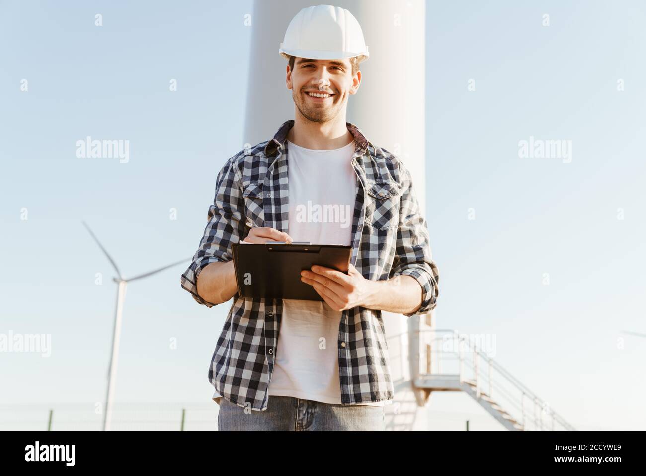 A smiling electrical engineer standing on a field with windmills ...