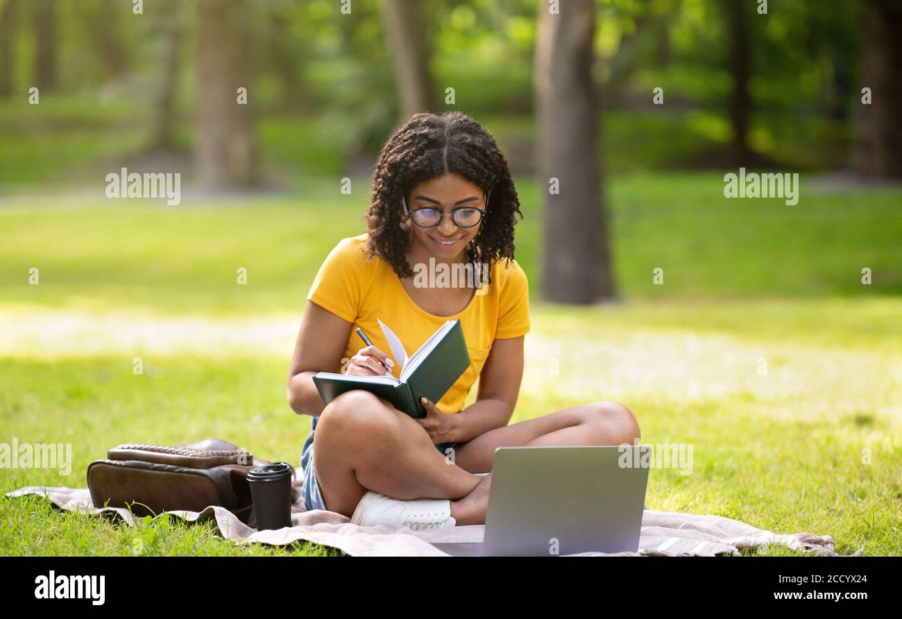 Focused millennial black girl writing something in notebook near laptop ...