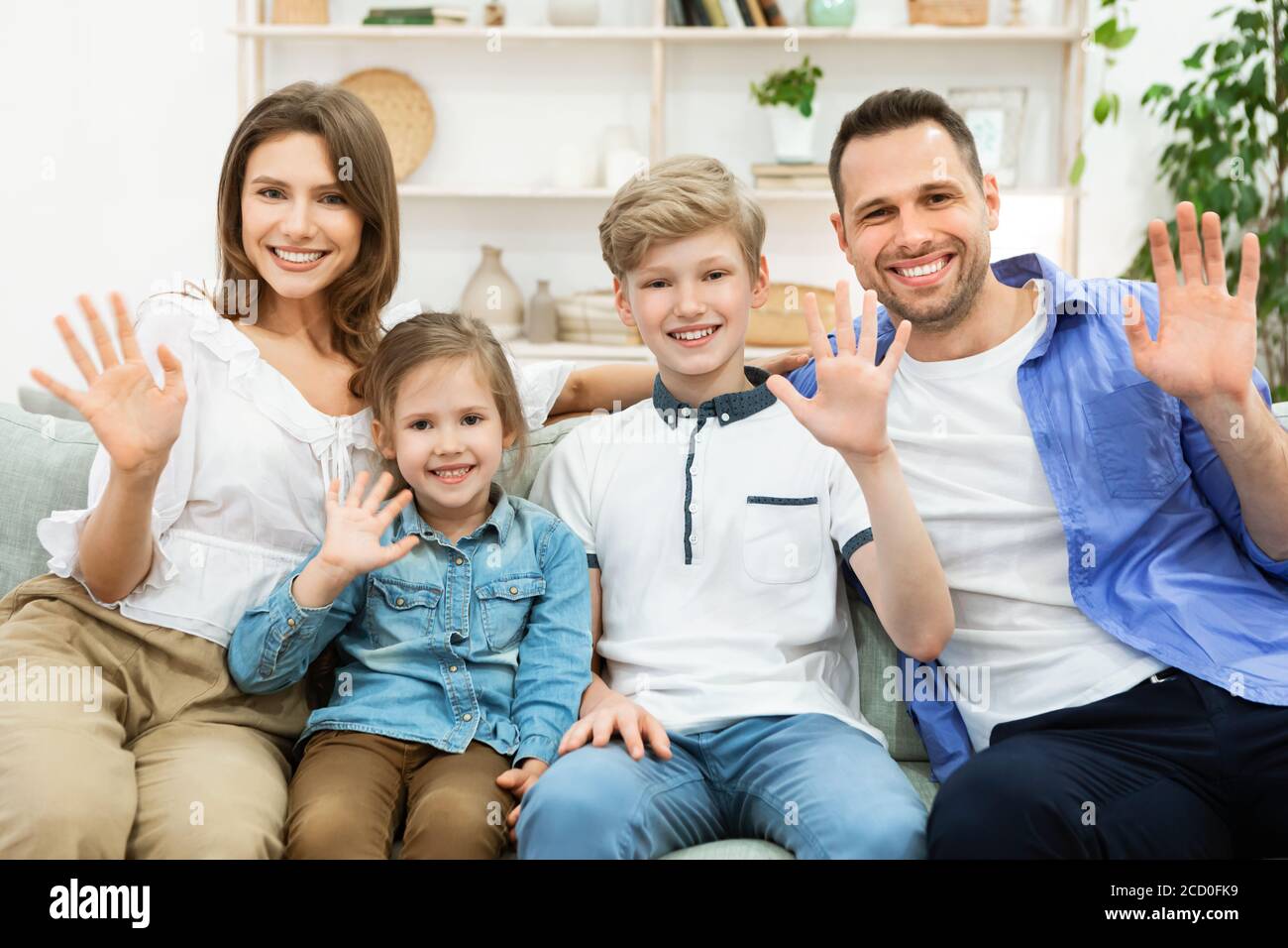 Family Of Four Waving Hello Greeting Sitting On Sofa Indoor Stock Photo ...