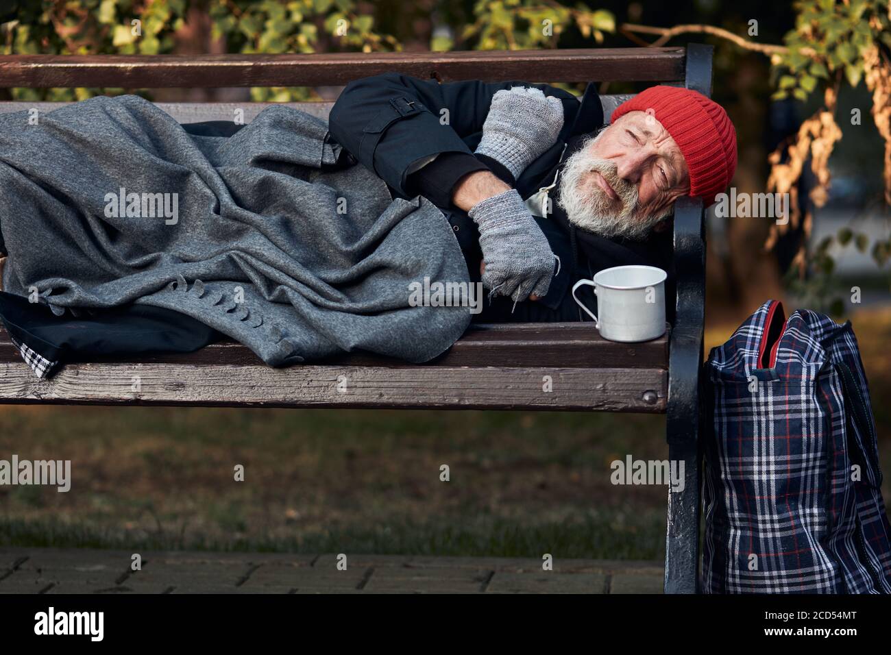 Penniless senior man lying on park bench, trembling from the cold. Cup ...