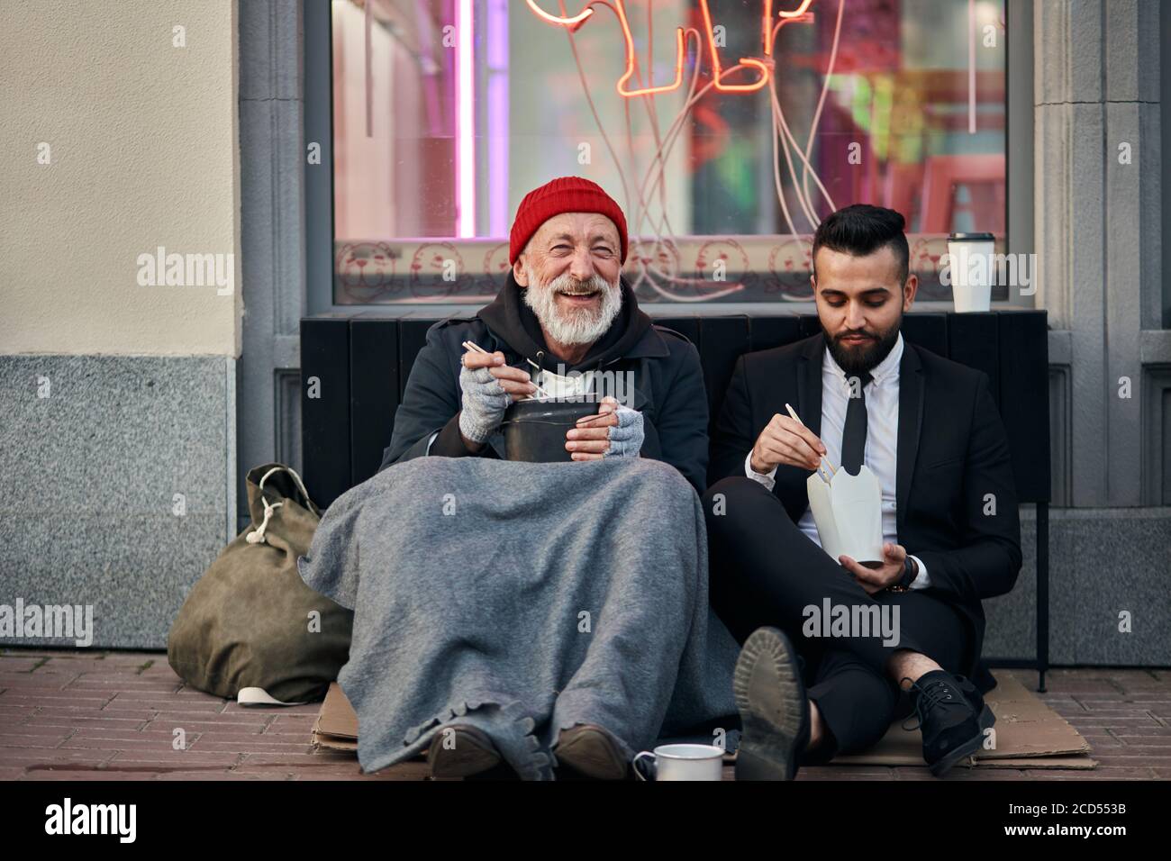 Smiling rich and poor men together sitting on street and eat while ...