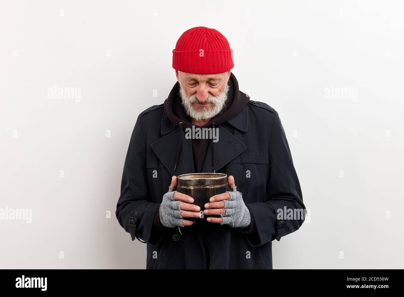 Sad old bum in black coat and red cap on head holding iron can for ...