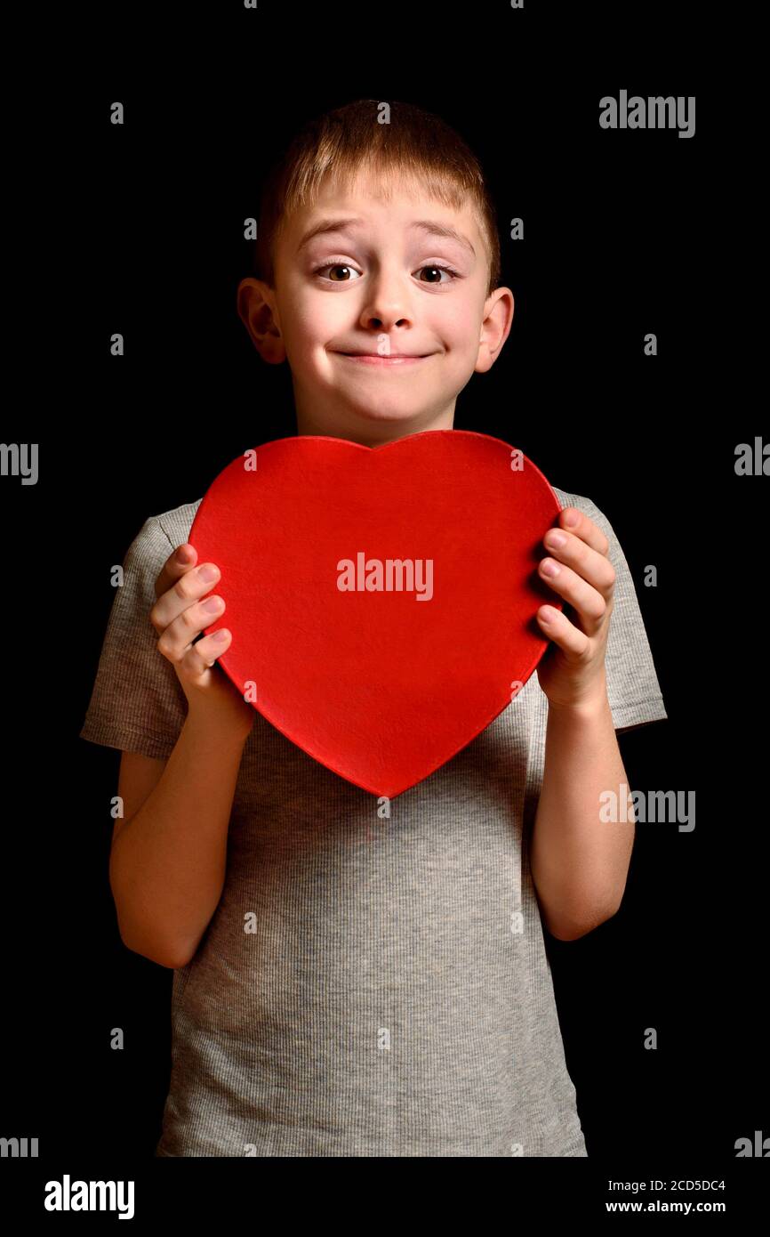 Blond boy holding a red box in the shape of a heart on black background ...