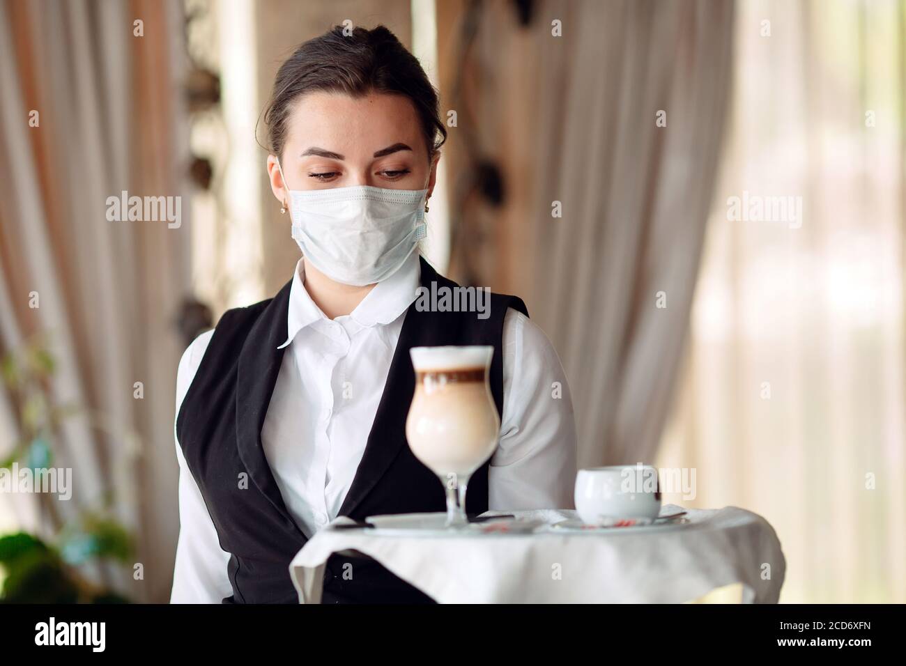 A female Waiter of European appearance in a medical mask serves Latte ...