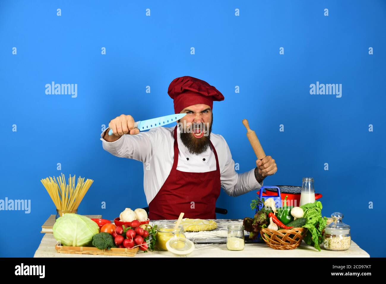 Chef with pasta, vegetables and dough on table. Cook with angry face in ...