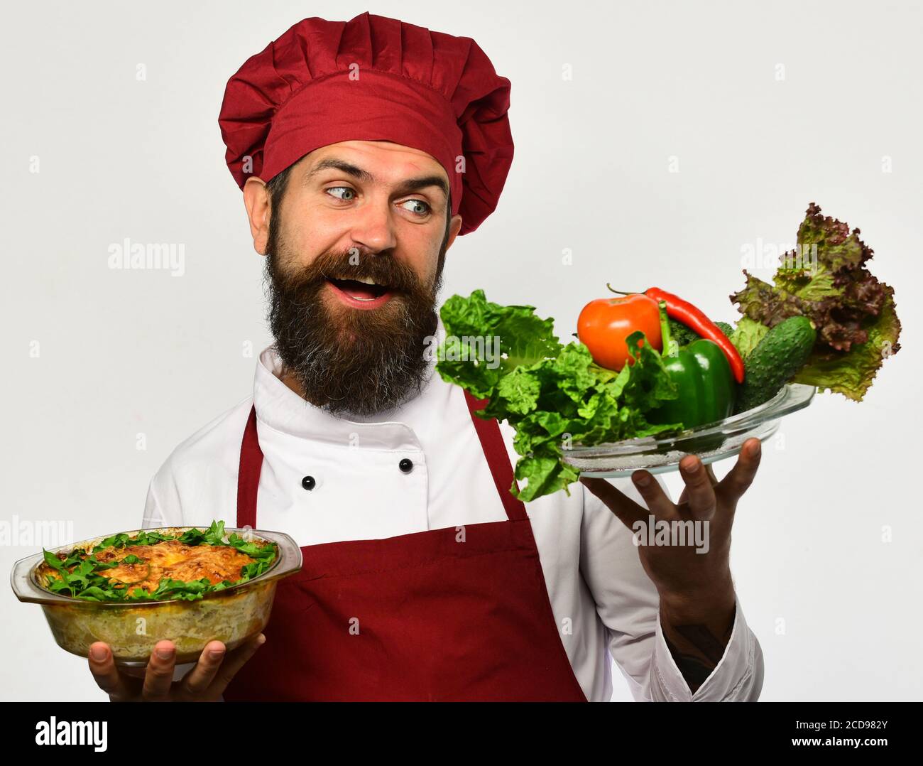 Cook with smile in burgundy uniform holds baked dish and salad. Cooking ...
