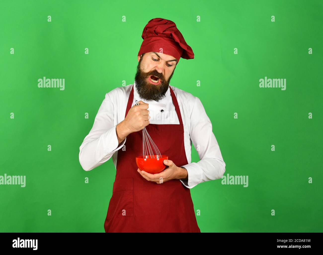 Cook with mad face in burgundy hat and apron holds bowl and whisk. Man ...