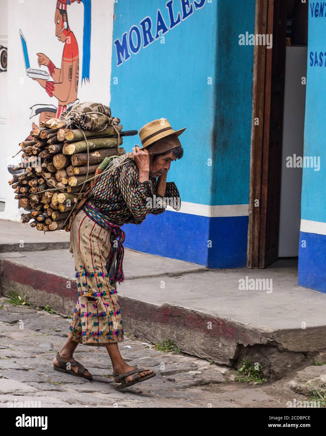 A poor Mayan man in ragged traditional clothes carries a heavy load of ...