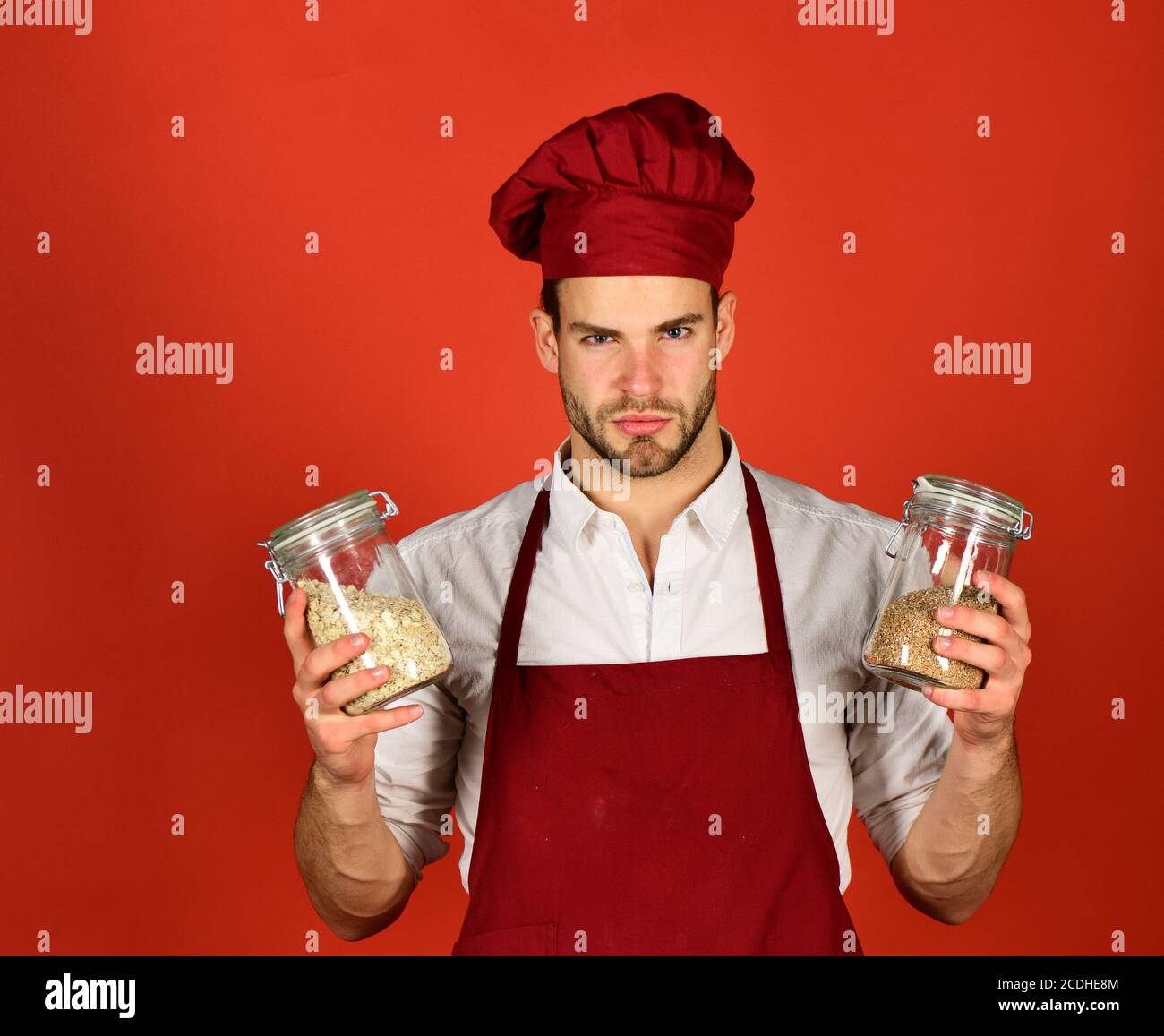 Chef with serious face holds jars with porridge on red background. Cook ...