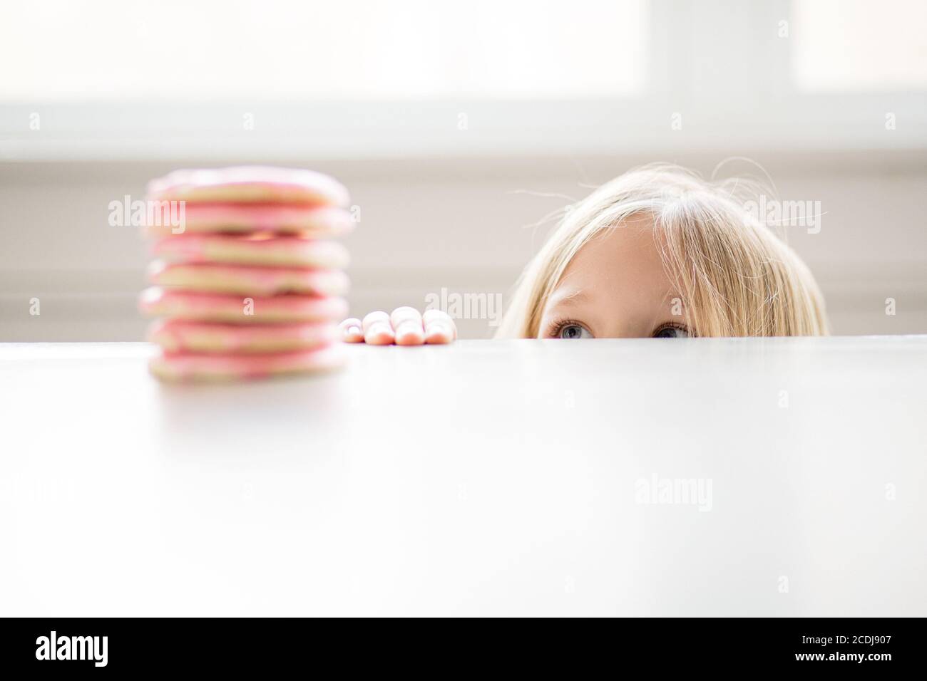 Blond girl peeking at stack of pink frosted sugar cookies on table ...