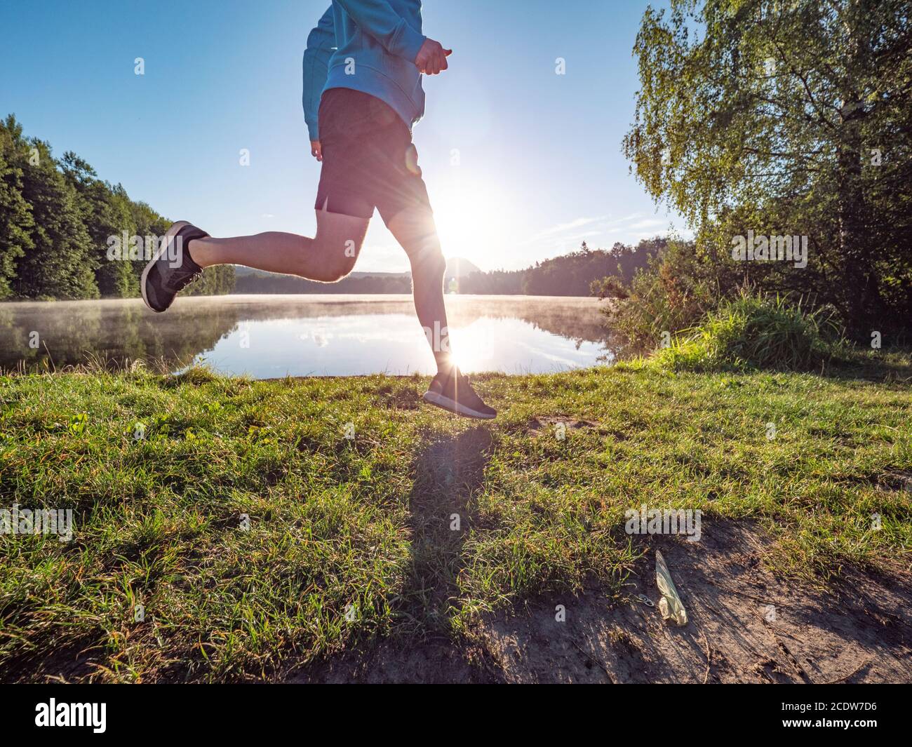 Athlete runner running on grassy beach at summer lake in rays of sunset ...