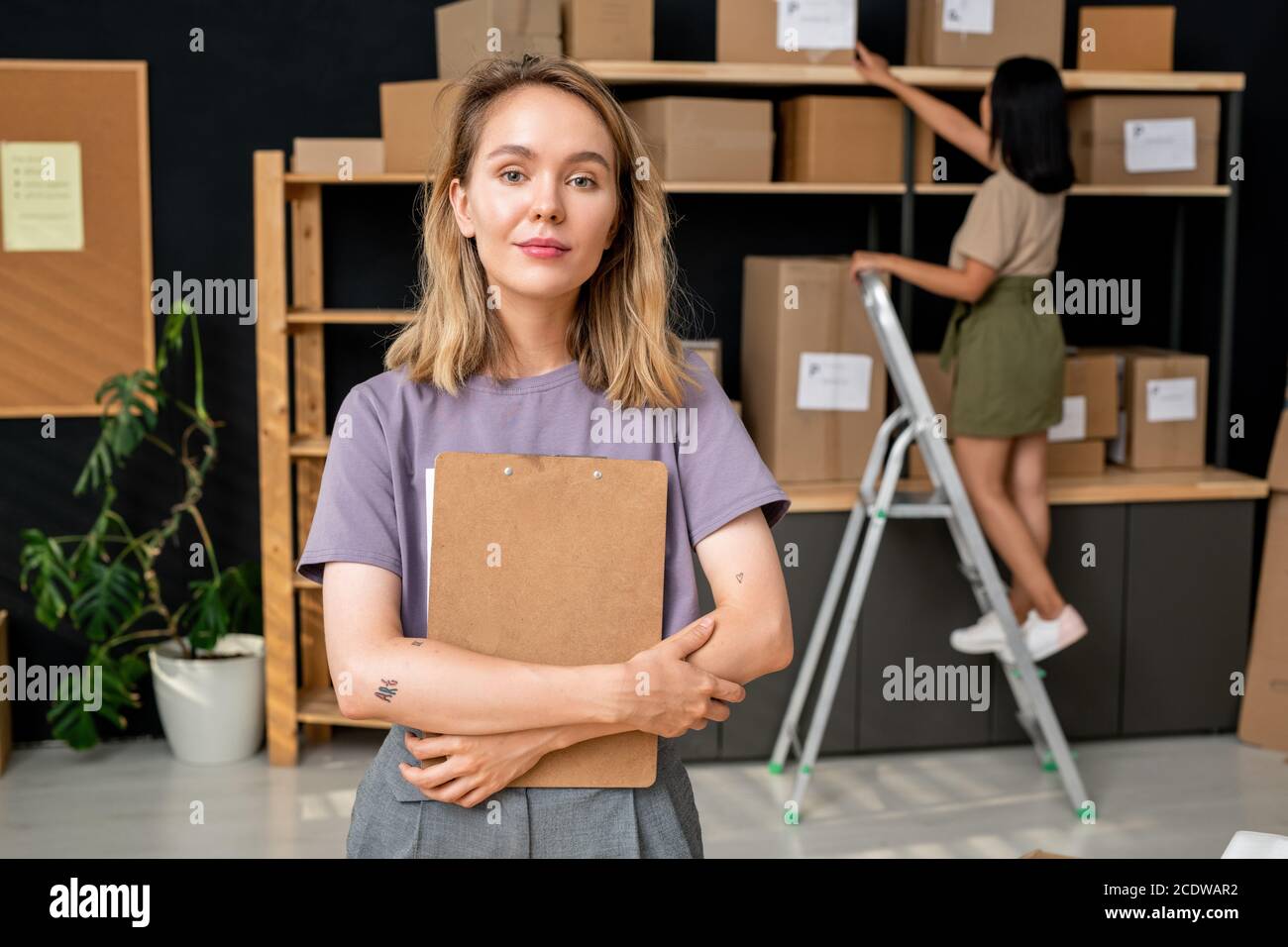 Young confident blond female worker of storage room with clipboard ...