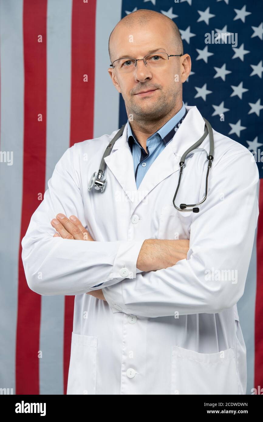 Bald doctor in whitecoat and eyeglasses against wall with American flag ...