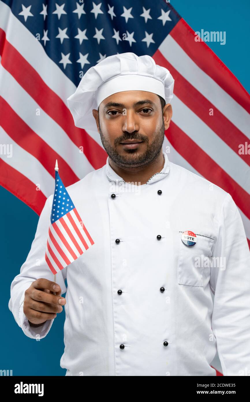Young serious chef in white uniform holding us flag against stars-and ...
