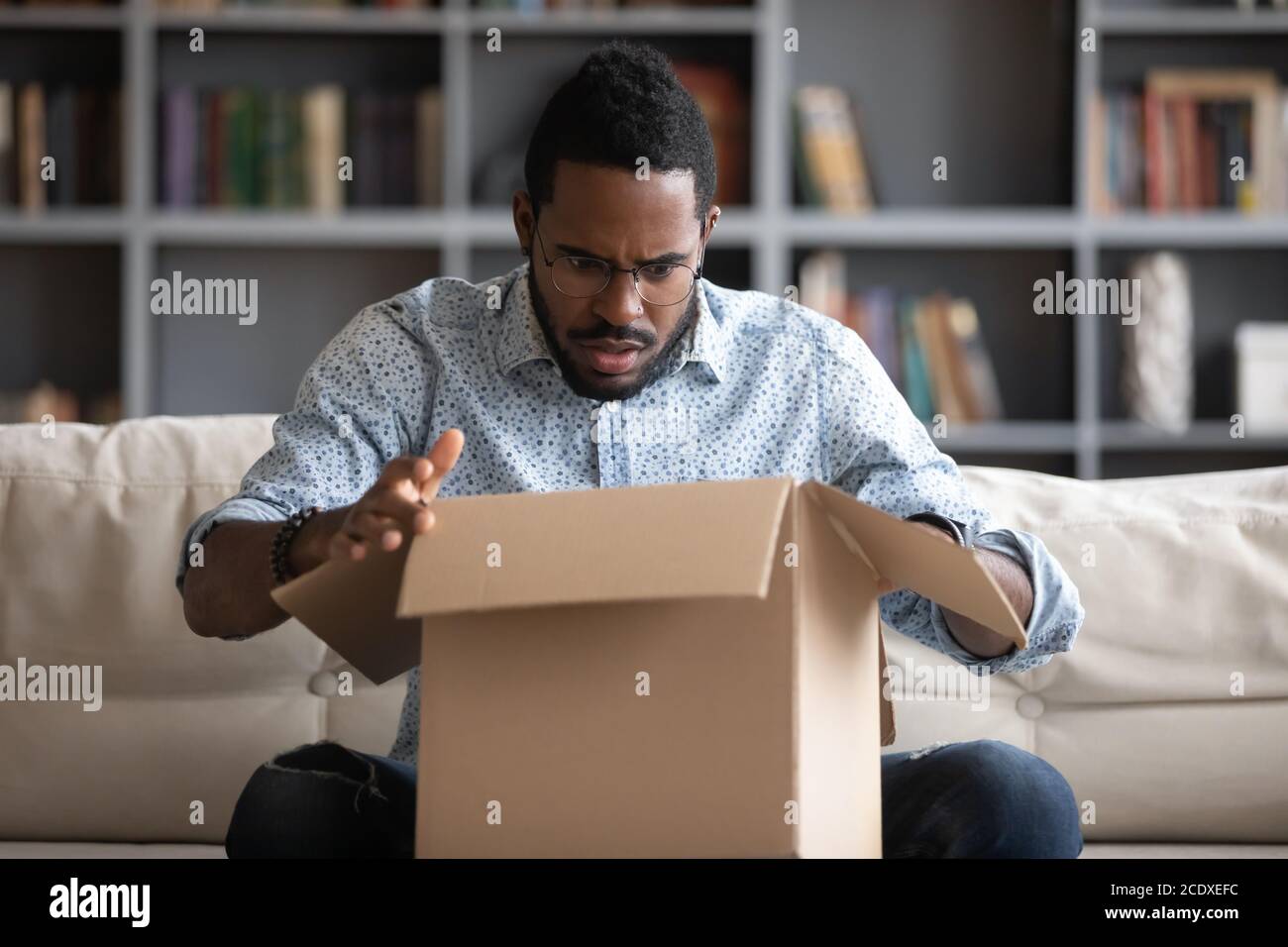 Unhappy shocked African American man unboxing parcel, looking inside ...