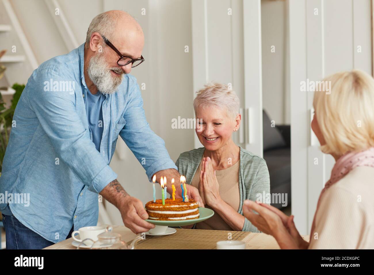 Happy senior woman sitting at the table while her friends bringing the ...