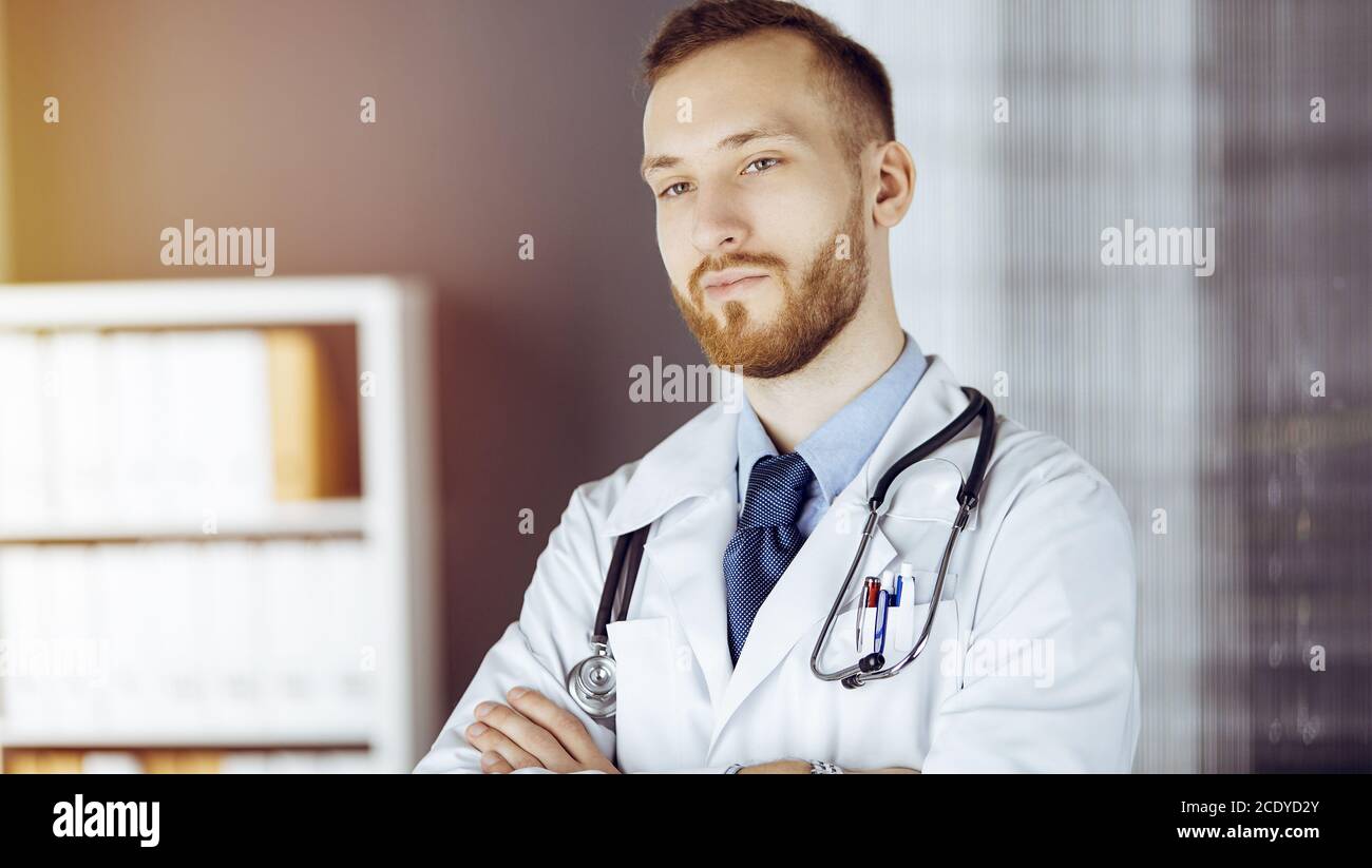 Red-bearded doctor standing straight in sunny clinic near his working ...