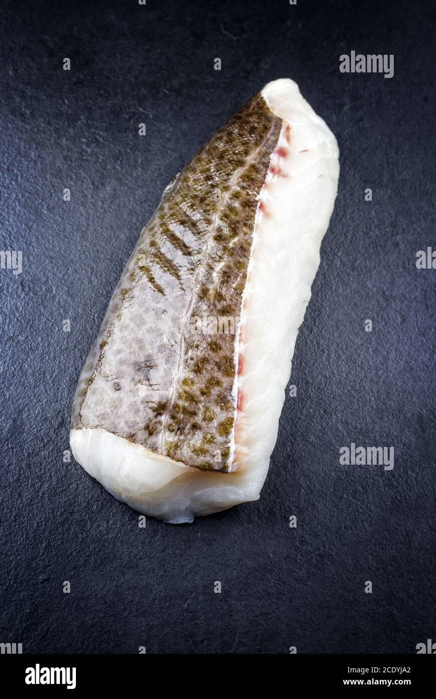 Raw Norwegian skrei cod fish filet with skin as closeup on black board ...