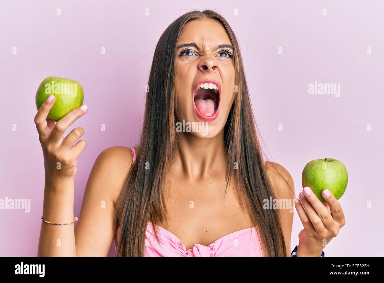 Young hispanic woman holding green apples angry and mad screaming ...