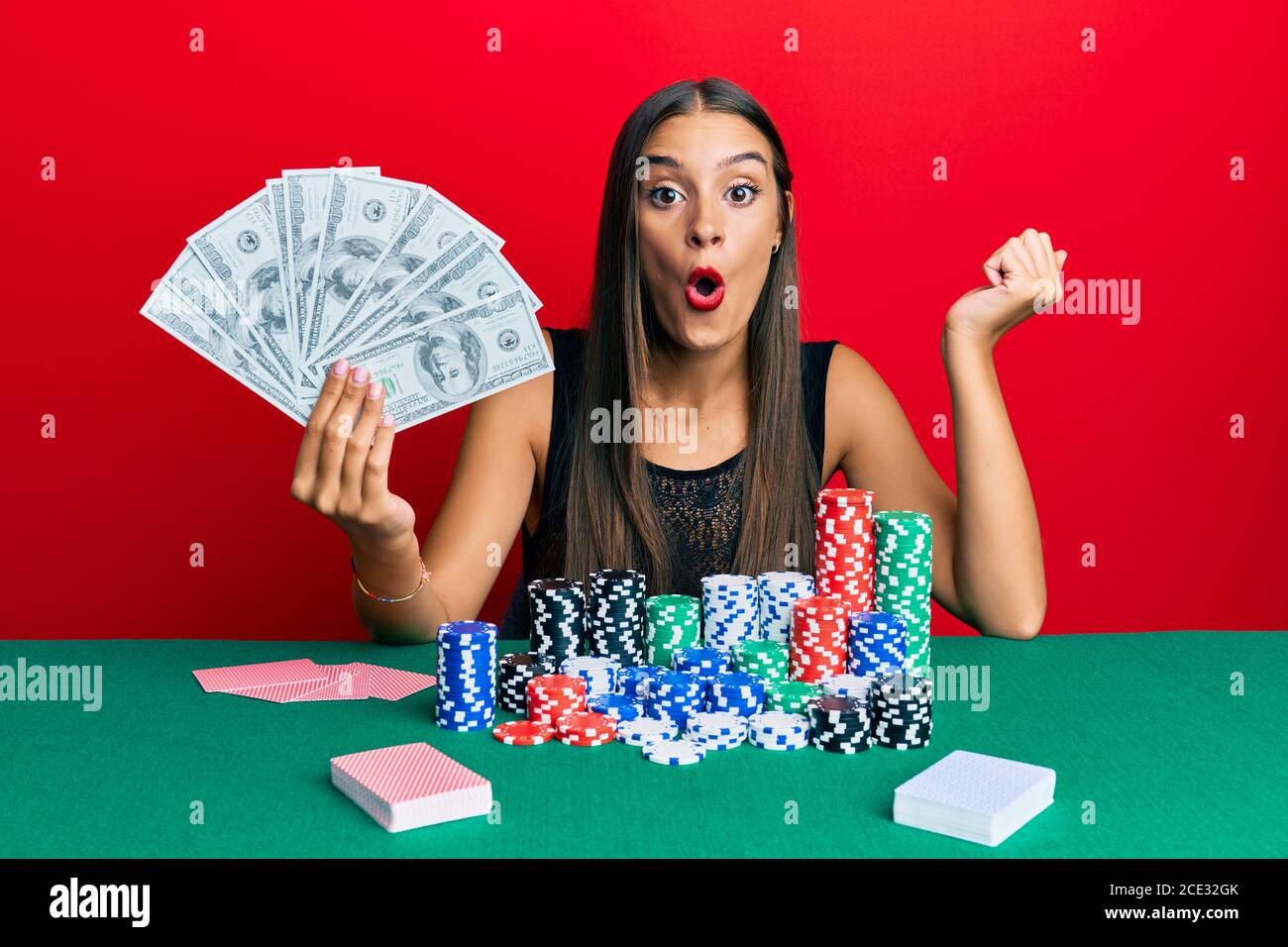Young hispanic woman playing poker holding dollars banknotes scared and ...