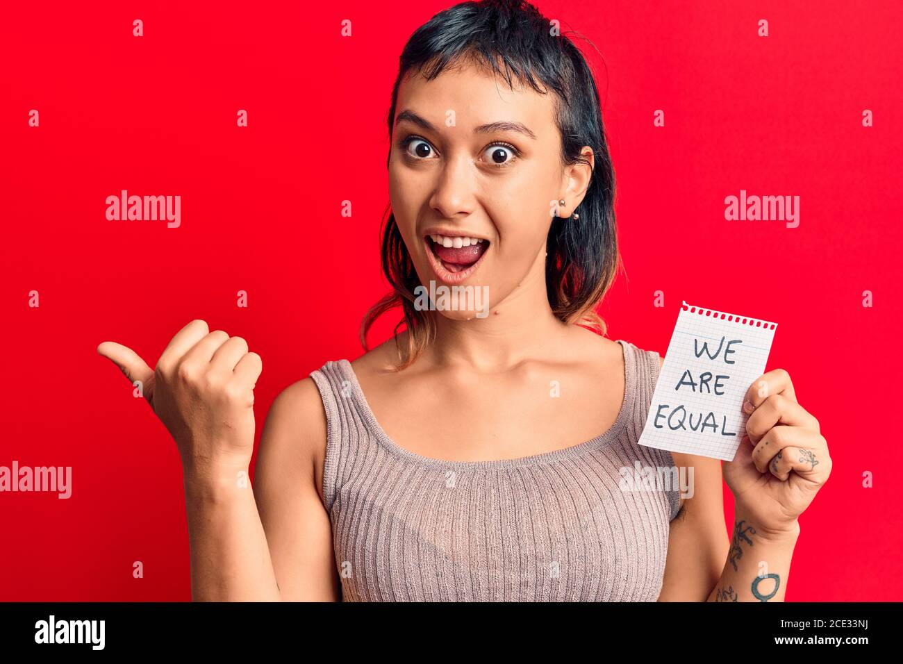 Young woman holding we are equal paper pointing thumb up to the side ...