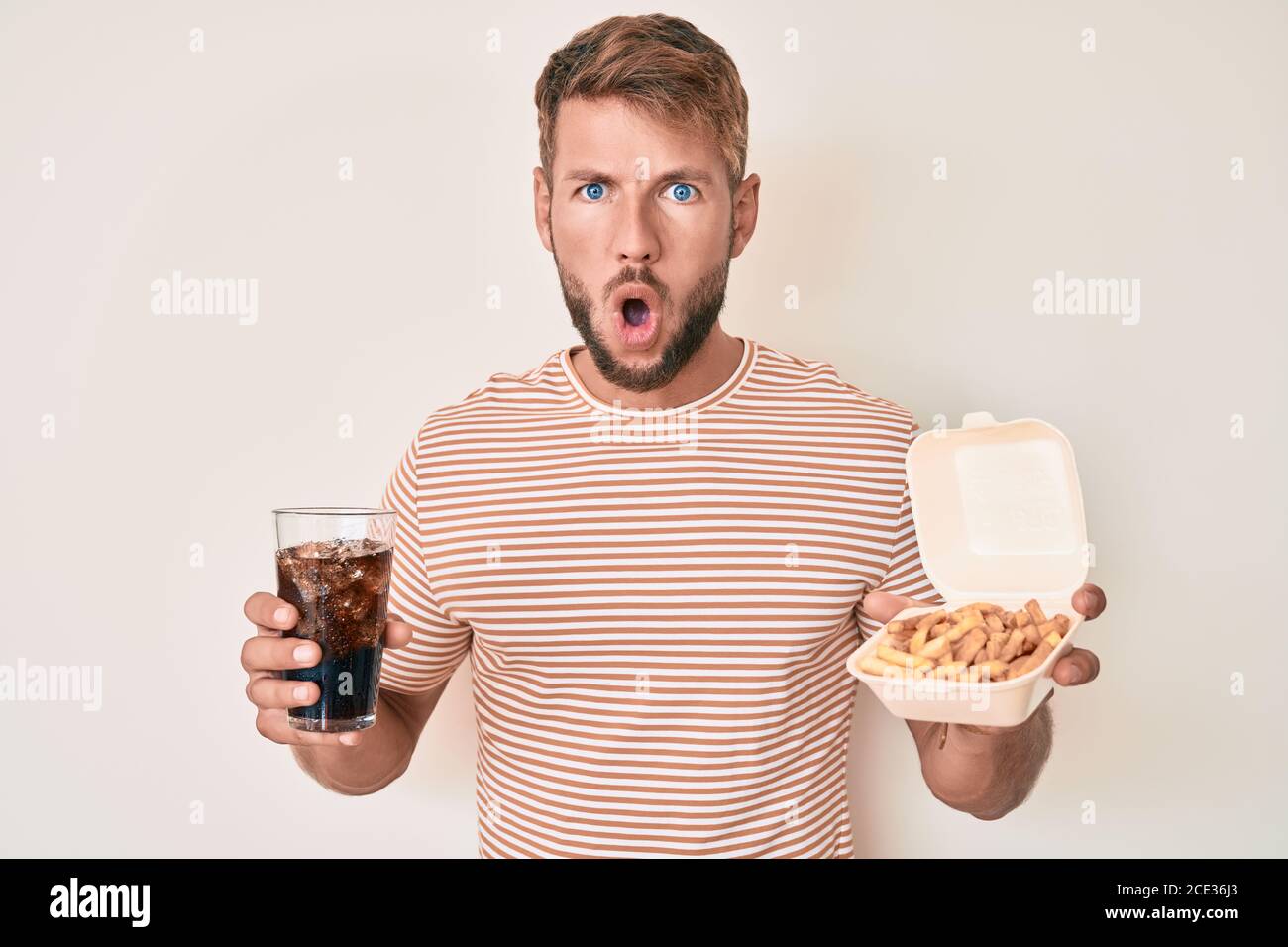 Young caucasian man eating fast food afraid and shocked with surprise ...