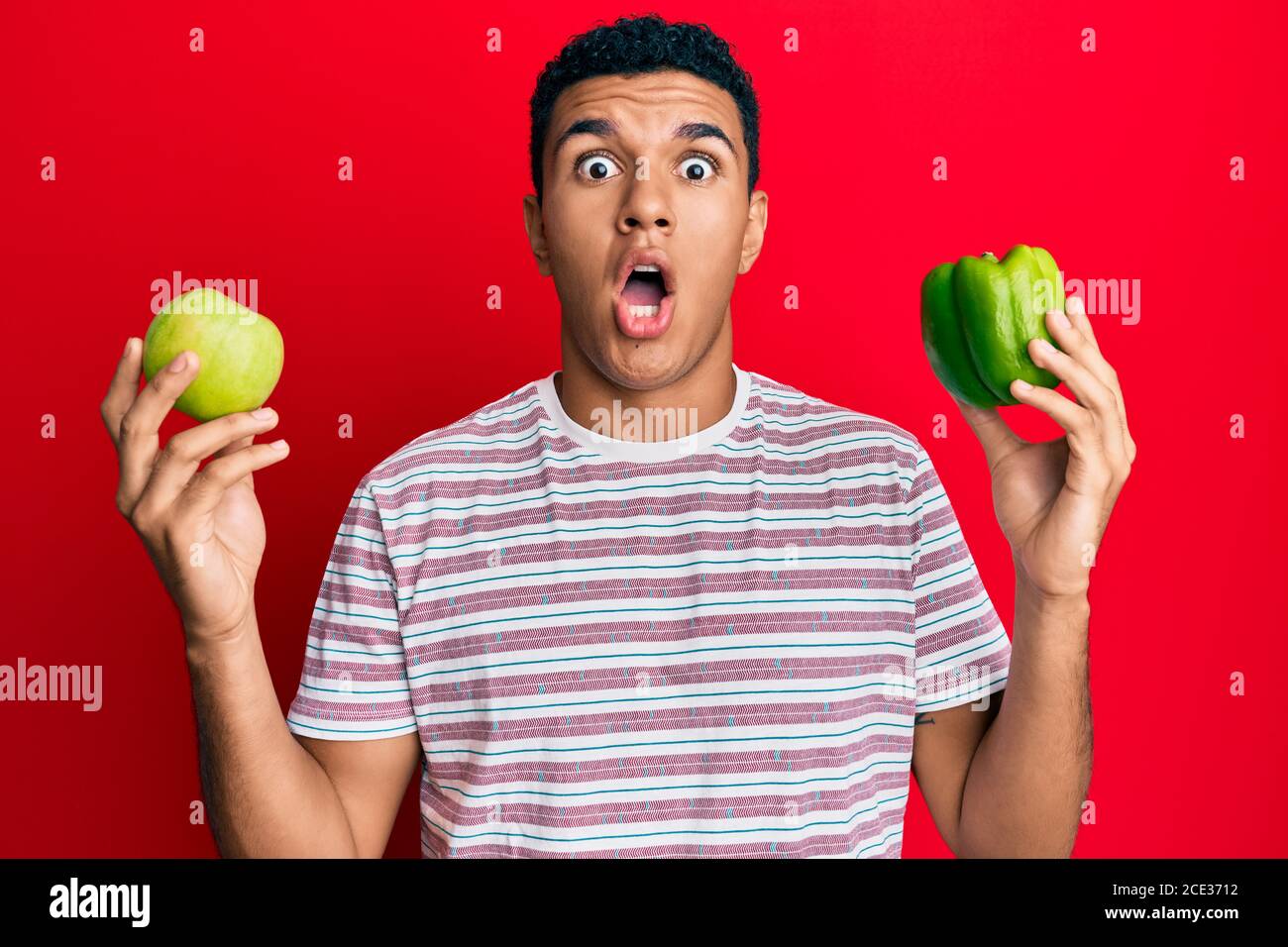 Young arab man holding green apple and pepper afraid and shocked with ...