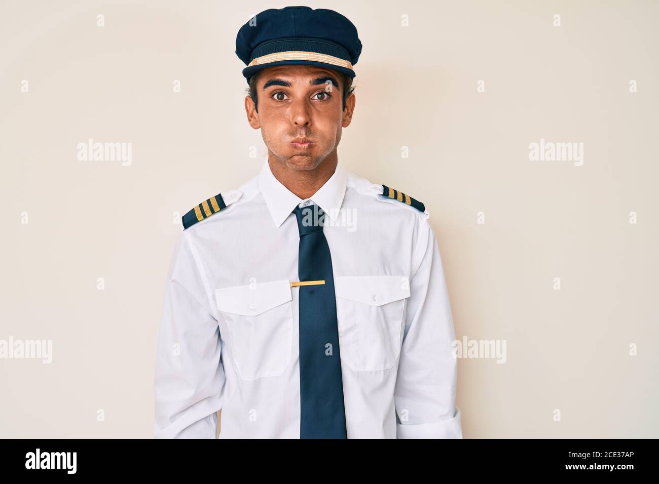 Young Hispanic Man Wearing Airplane Pilot Uniform Puffing Cheeks With
