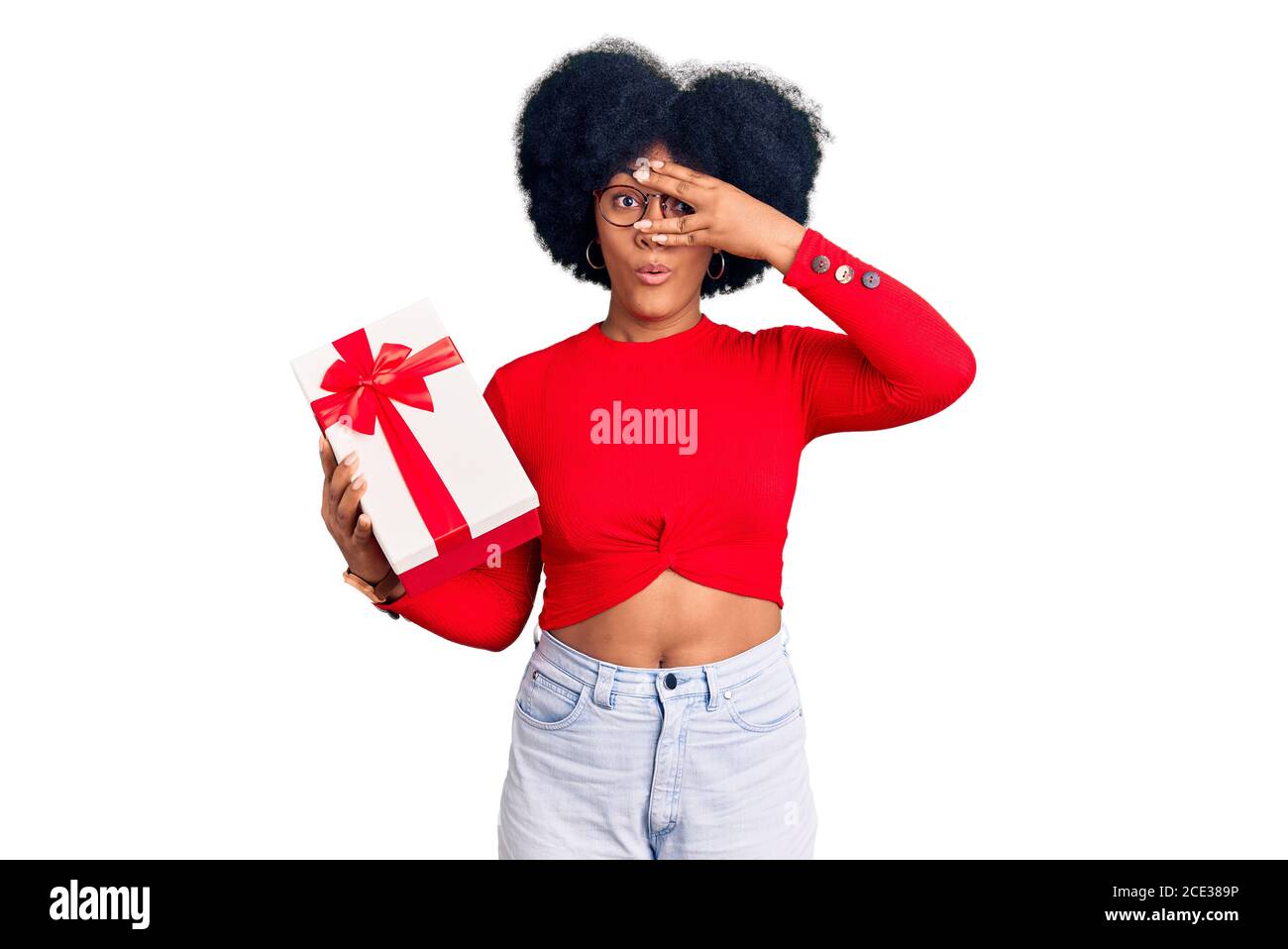 Young african american girl holding gift peeking in shock covering face ...